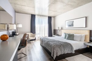 Quarto de hotel moderno com uma cama grande, uma escrivaninha com abajur, uma cadeira e vista para a cidade pela janela.
