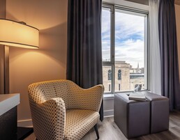 Quarto de hotel moderno com uma poltrona estampada, um pequeno banquinho, uma luminária e uma janela ampla com vista para a cidade.