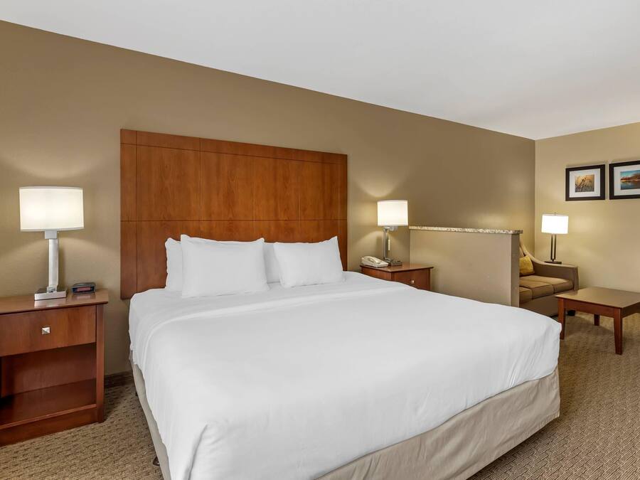 Quarto de hotel com uma cama grande, duas mesas de cabeceira com luminárias, uma cabeceira de madeira, um sofá pequeno e duas fotos emolduradas na parede.