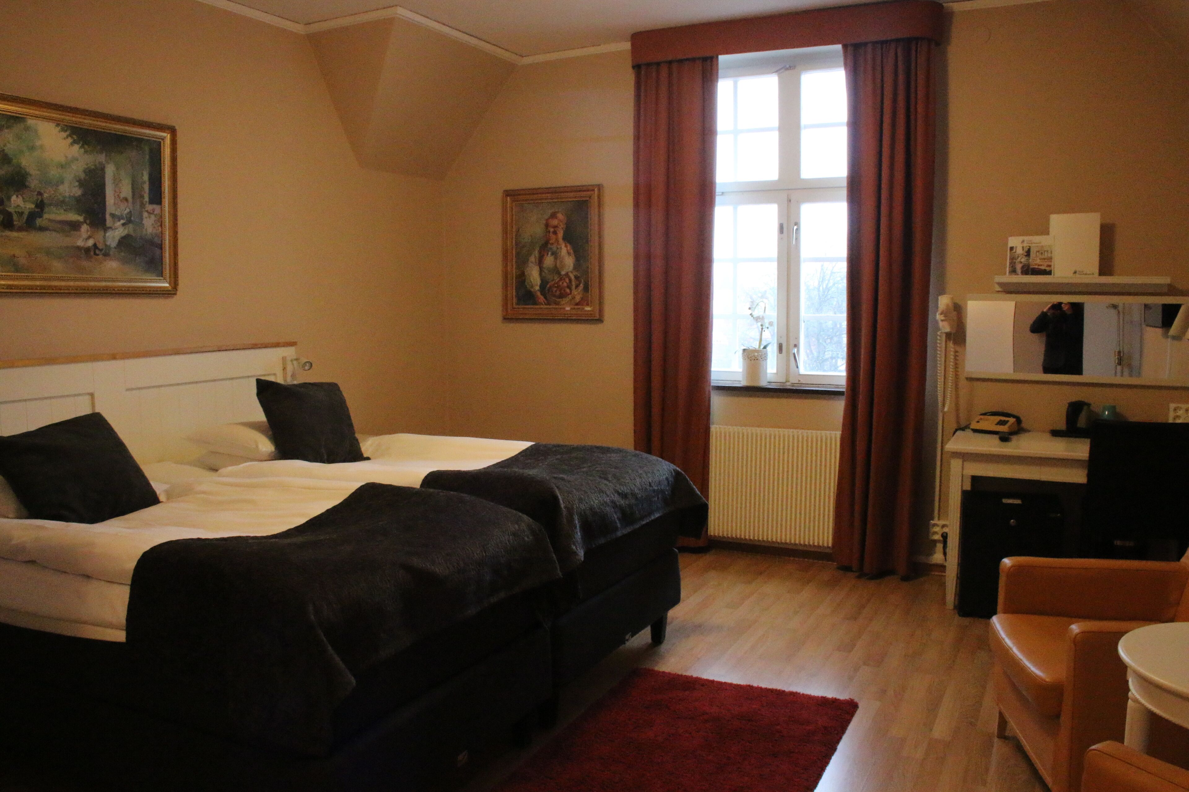 Quarto de hotel com duas camas, uma mesa, uma cadeira, um radiador e uma janela com cortinas.