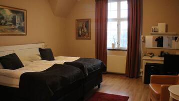 Quarto de hotel com duas camas, uma mesa, uma cadeira, um radiador e uma janela com cortinas.
