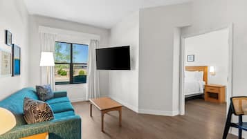 Quarto de hotel com sofá, mesa pequena, cama, televisão e janela com cortinas.