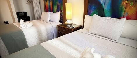 Quarto de hotel com duas camas, uma mesa de cabeceira com abajur, um quadro na parede e um cabideiro.