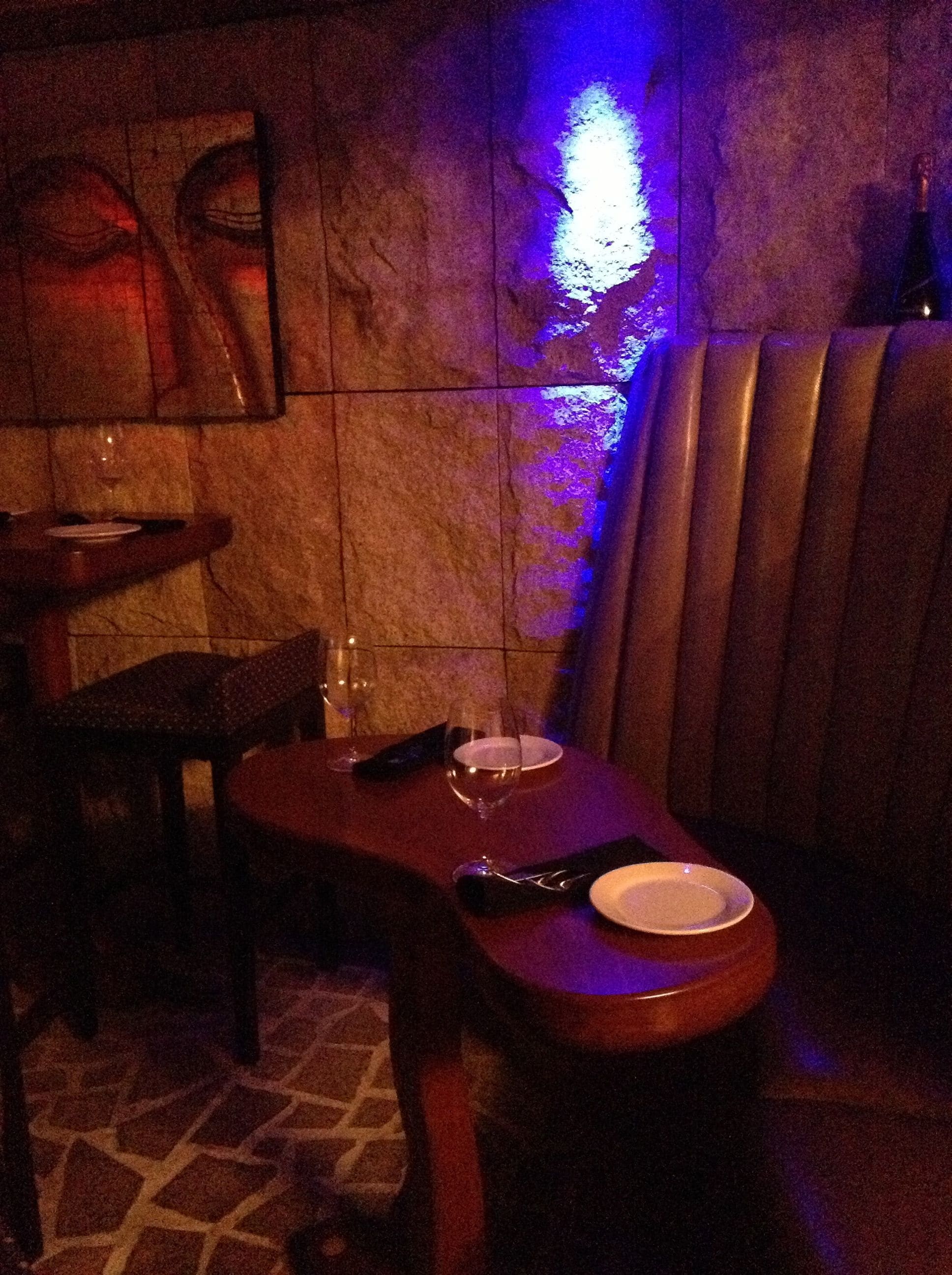 Restaurante com iluminação fraca, parede de mármore, mesa de madeira com um prato e um copo, e uma cadeira.