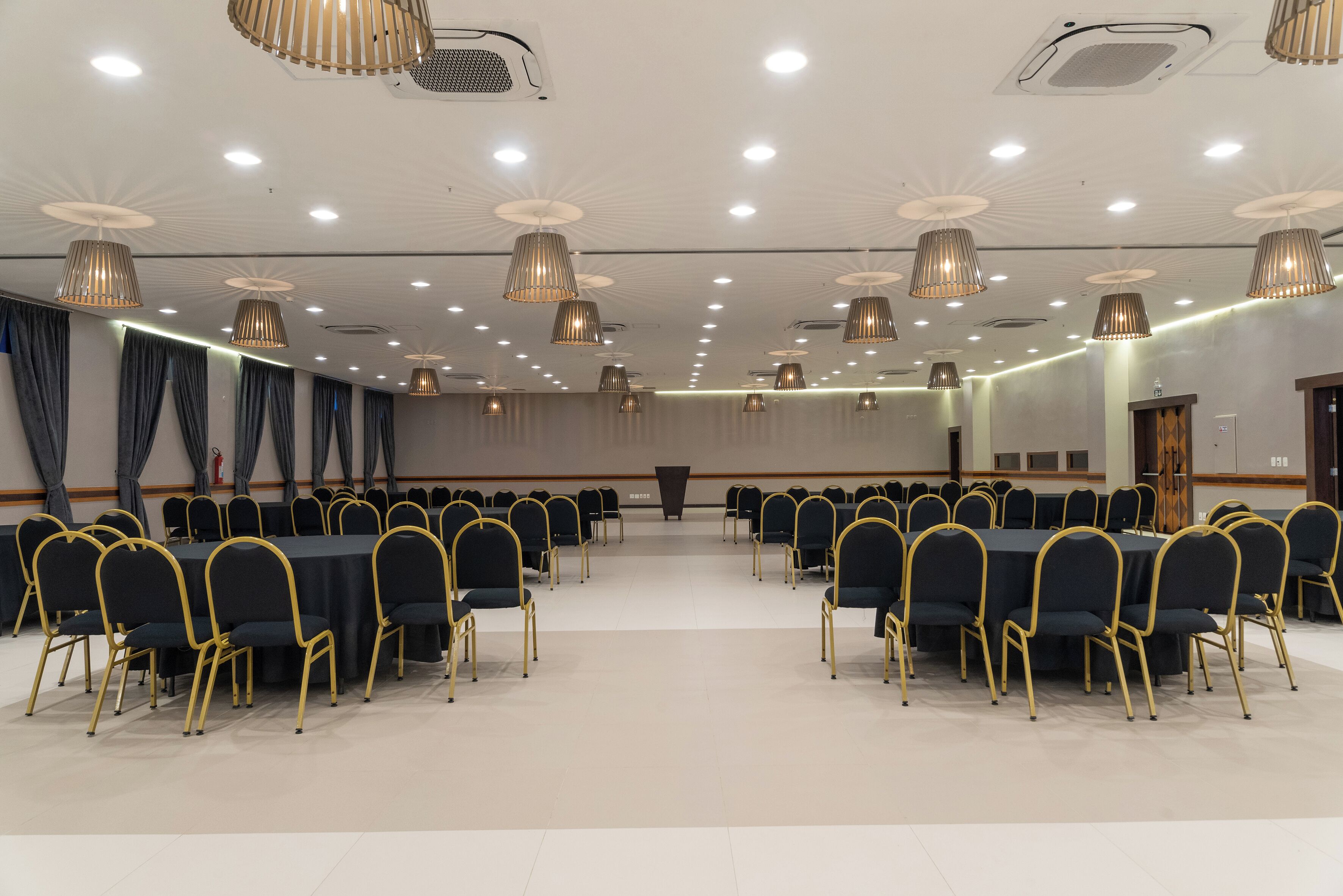 Uma sala de conferências com mesas e cadeiras dispostas em fileiras, luminárias embutidas no teto e lustres pendentes.