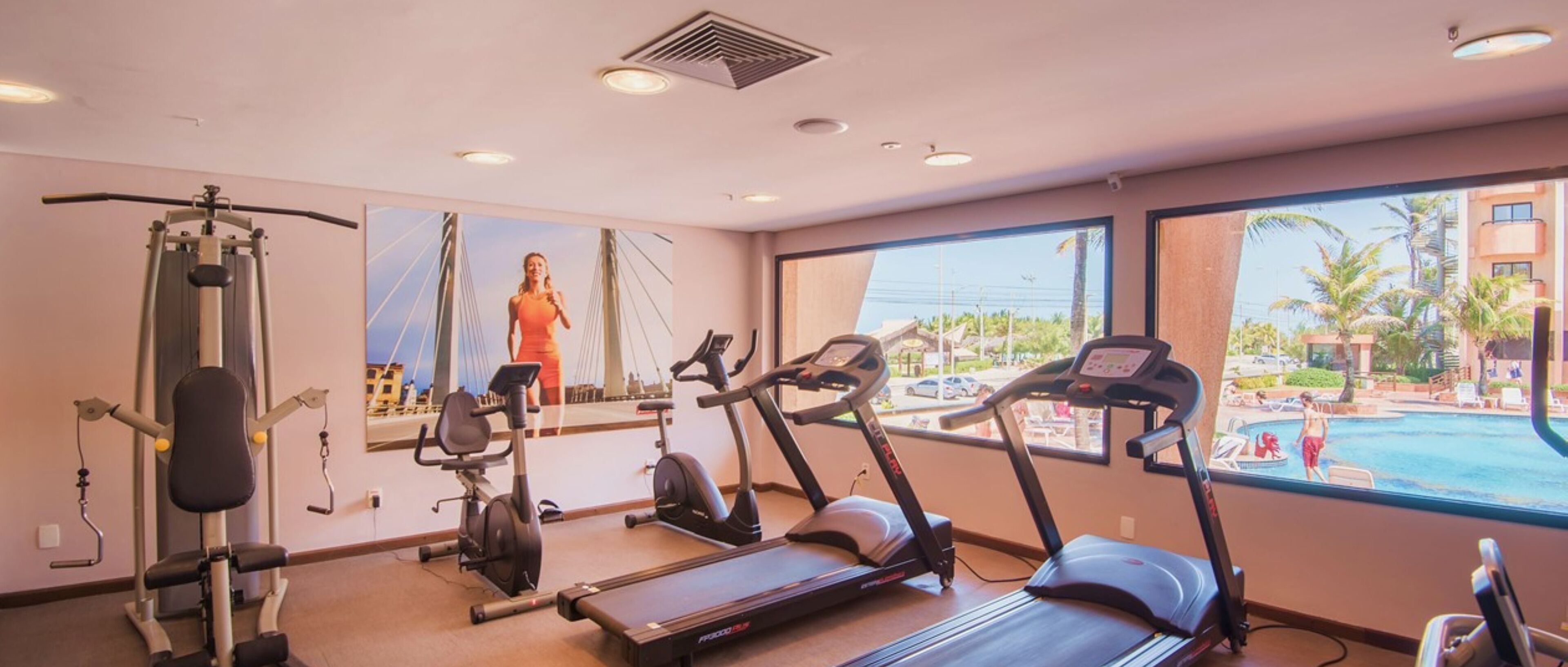Uma academia com equipamentos de ginástica, uma janela ampla com vista para uma piscina e palmeiras.
