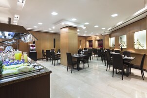 Espaço de refeições de hotel com várias mesas e cadeiras, buffet com diversos alimentos e bebidas, e design de interiores moderno.