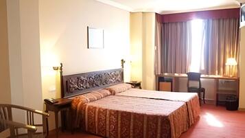 Quarto de hotel com uma cama grande, uma escrivaninha, uma cadeira e uma janela com cortinas.