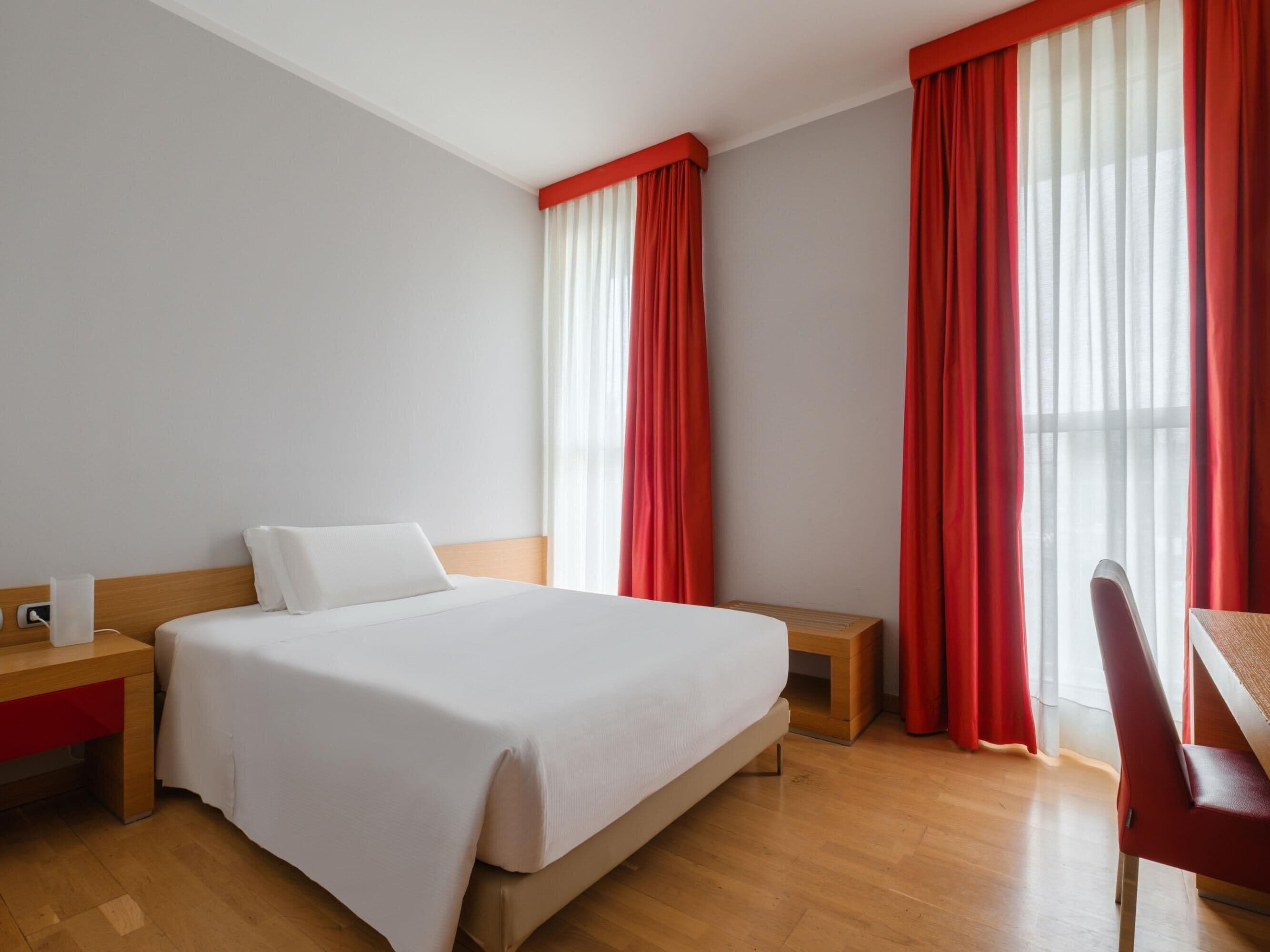 Quarto de hotel com cama, mesas de cabeceira, uma cadeira e cortinas vermelhas.