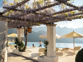 Área de piscina com espreguiçadeiras, guarda-sóis e um pergolado com flores roxas.