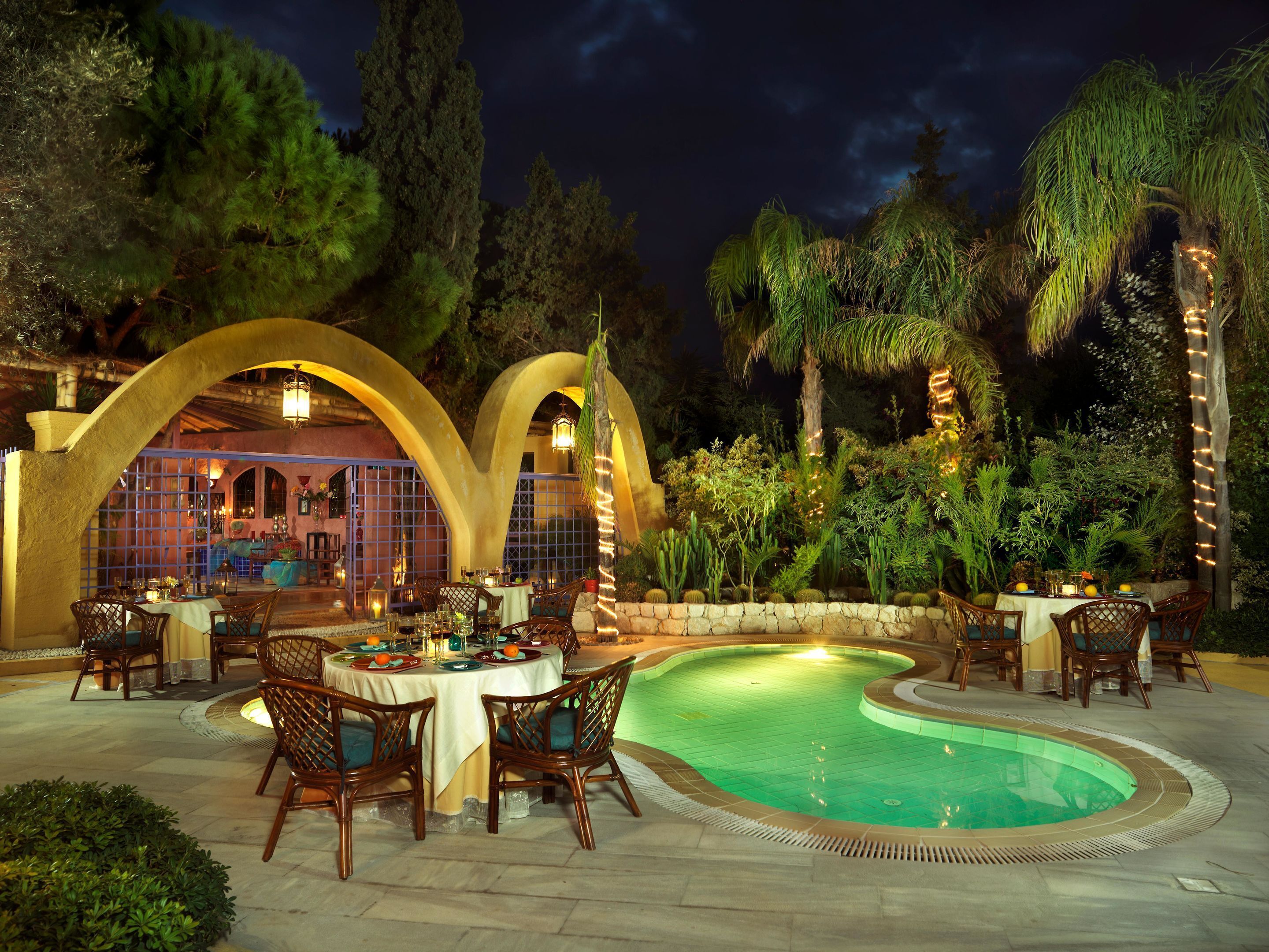 Uma área de refeições ao ar livre com mesas e cadeiras, uma piscina e um terraço com plantas e árvores.