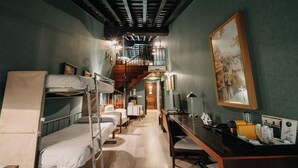 Quarto de hotel com beliches, uma escrivaninha, uma cadeira, máquina de café e escada que leva a um andar superior.