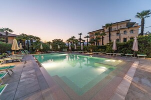 Uma piscina com espreguiçadeiras e guarda-sóis, rodeada por vegetação e um edifício com varandas.