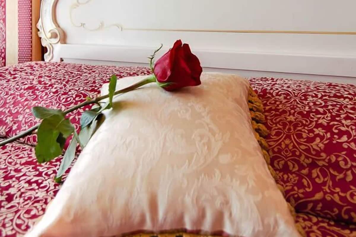 Uma cama com cabeceira branca e ornamentada, um edredom estampado em vermelho e dourado, e uma única rosa vermelha sobre o travesseiro.