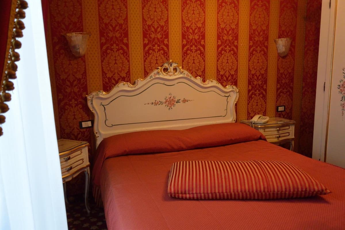 Cama com cabeceira branca e roupa de cama vermelha em quarto com papel de parede listrado.