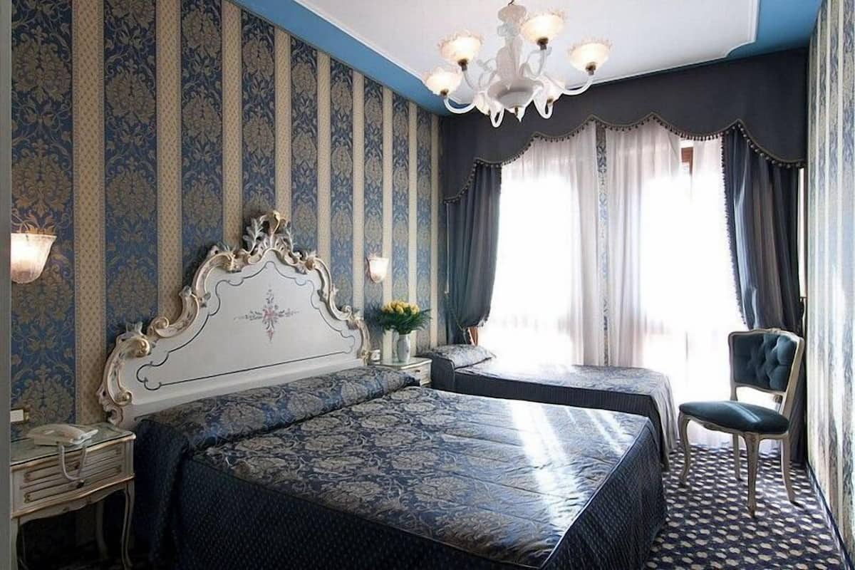 Quarto de hotel com uma cama grande, uma cama menor, uma escrivaninha com abajur e uma cadeira.