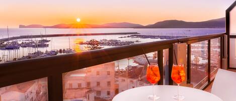 Varanda com vista para uma marina e pôr do sol. Dois copos com bebida vermelha estão sobre uma mesa.