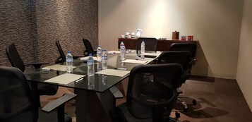 Sala de conferência com mesa de vidro, cadeiras pretas, garrafas de água e um canto para café.
