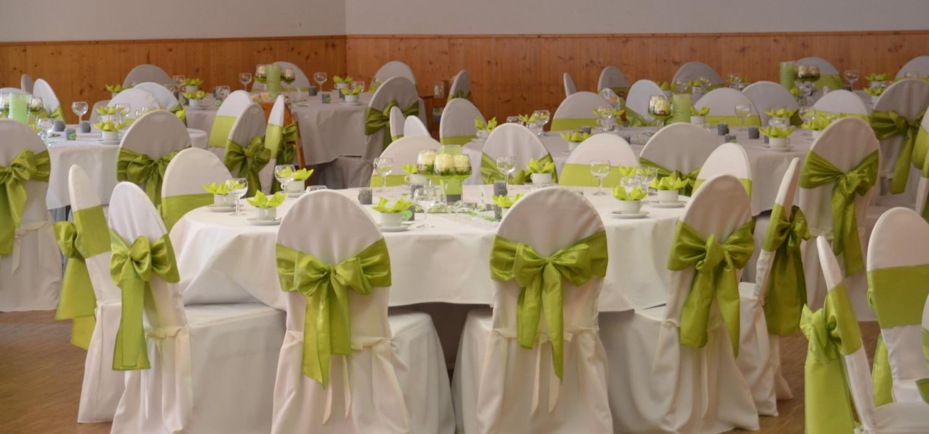 Uma sala de banquetes com mesas redondas cobertas por toalhas brancas, cada uma rodeada por cadeiras com fitas verdes.