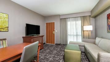 Quarto de hotel com televisão de tela plana, uma escrivaninha de madeira, um banquinho verde, um sofá bege e uma janela grande com cortinas.