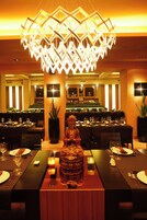 Um restaurante com um lustre central, várias mesas de jantar e um bar ao fundo.