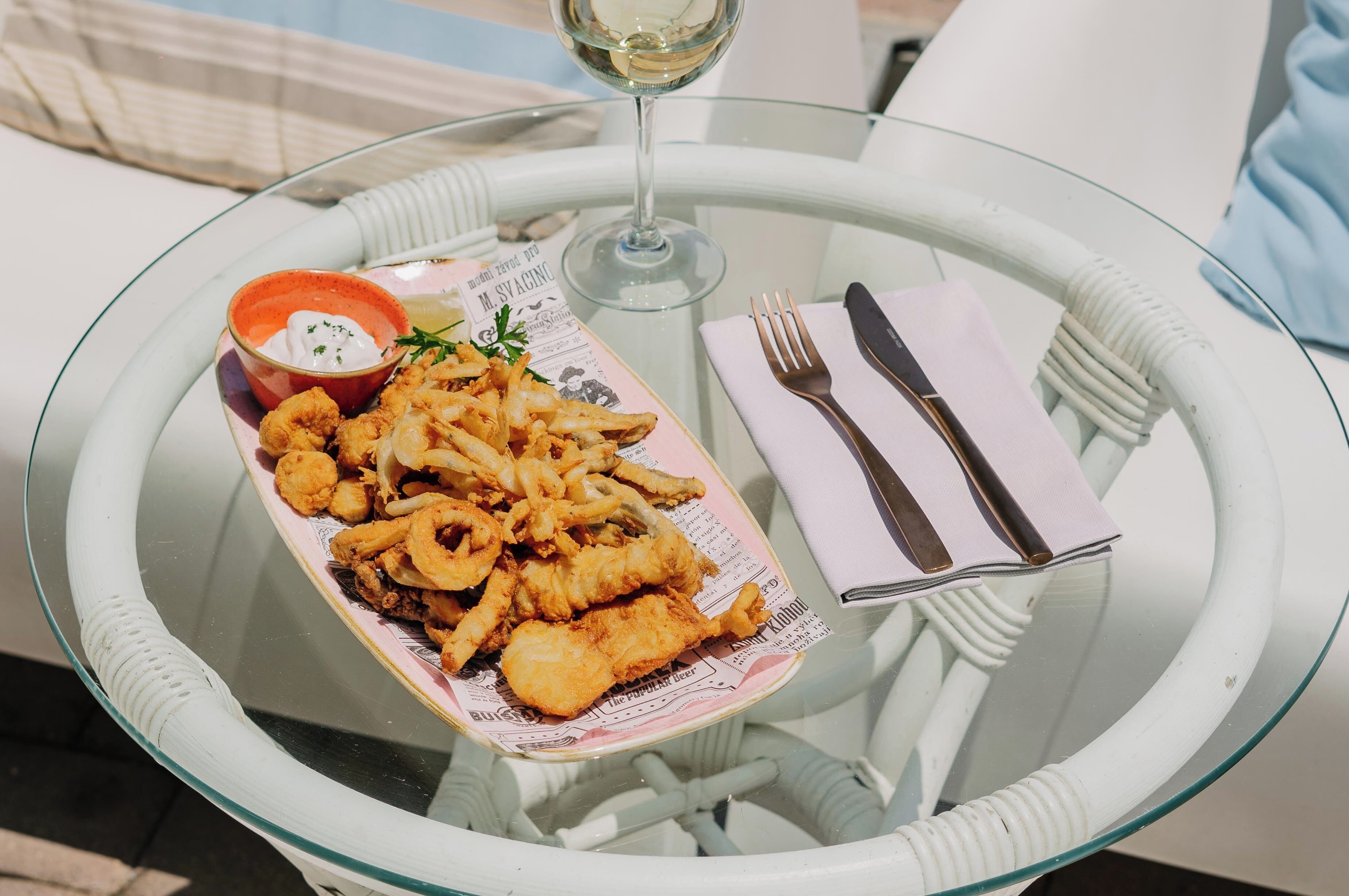 Uma mesa de vidro com um prato de frutos do mar fritos, uma pequena tigela de molho e um copo de vinho branco.