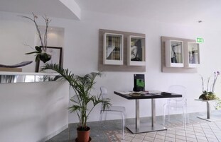 Uma área de recepção com balcão, duas fotos emolduradas na parede, um vaso de plantas e uma mesa de vidro com cadeiras.