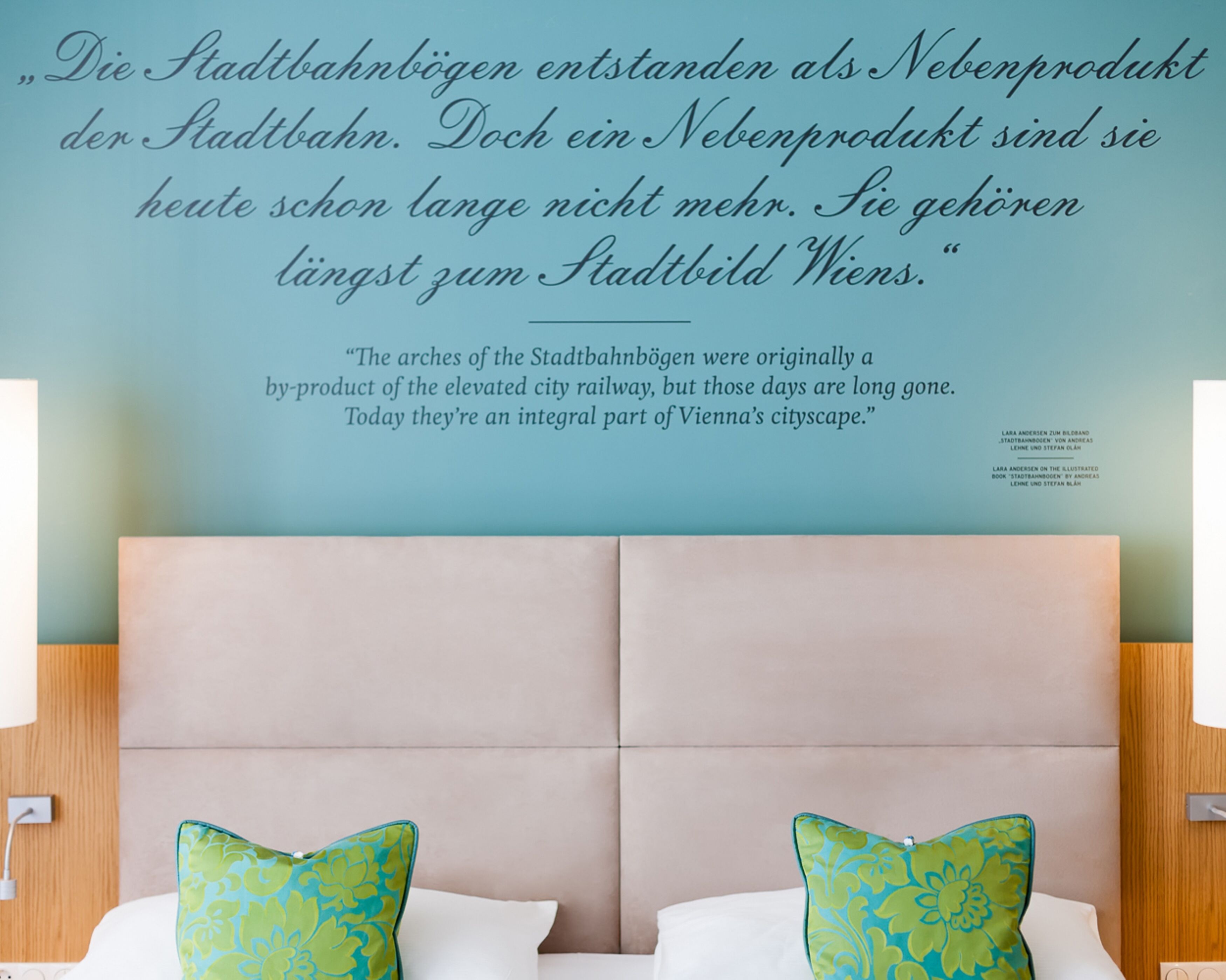 Quarto de hotel com uma cama grande, duas almofadas decorativas e uma parede com uma citação em alemão.