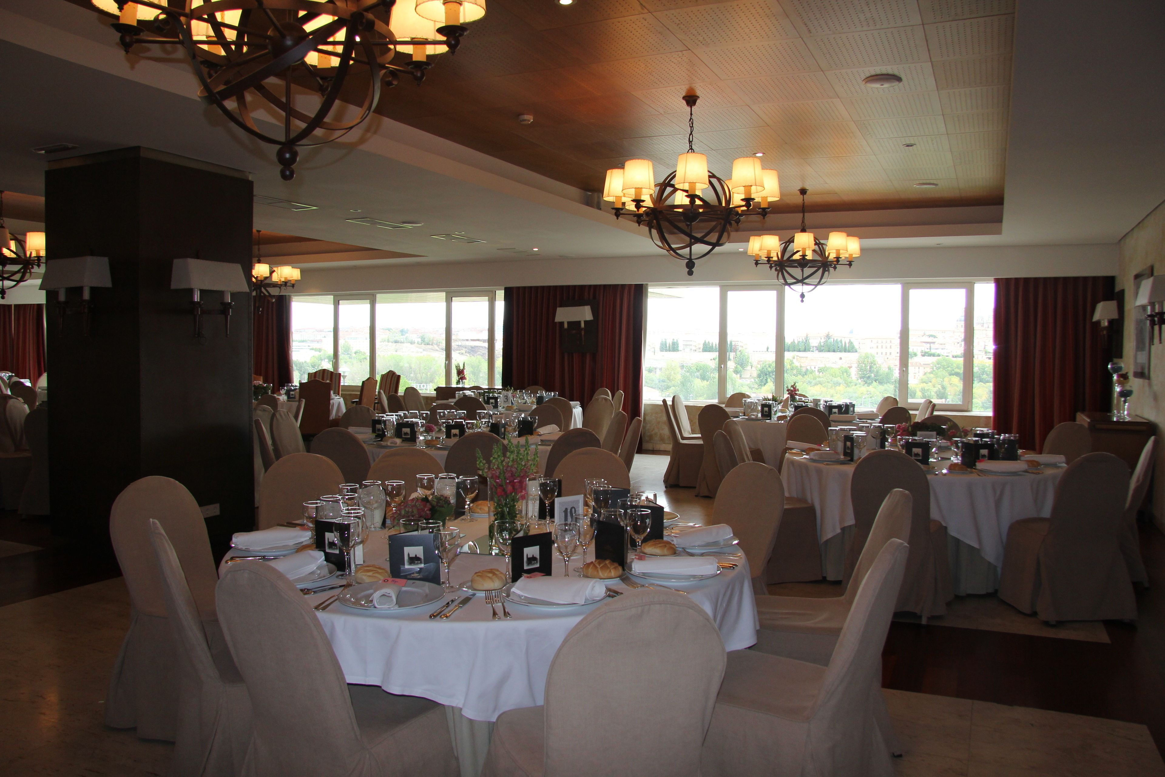 Salão de banquetes com várias mesas redondas preparadas para refeições, amplas janelas com vista para o exterior e lustres ornamentados.