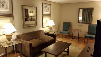 Uma sala de estar mobiliada com sofá, mesa de centro, poltrona e televisão. Há quadros nas paredes e um abajur sobre uma mesinha lateral.