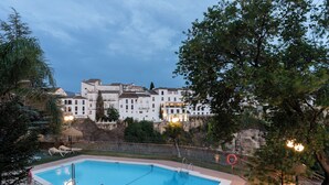 Piscina com espreguiçadeiras, um telhado de palha e vista para edifícios brancos ao fundo.