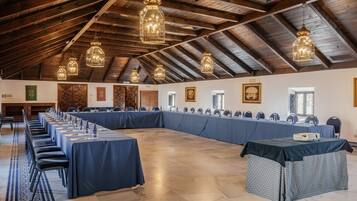 Uma longa mesa de conferências com toalhas azuis e cadeiras, num espaço com vigas de madeira e luminárias pendentes estilo lanterna.