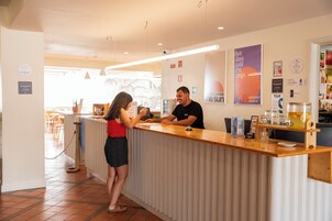 Uma área de recepção com balcão de madeira, uma mulher e um homem atrás dele, e diversos serviços como uma máquina de café e copos.
