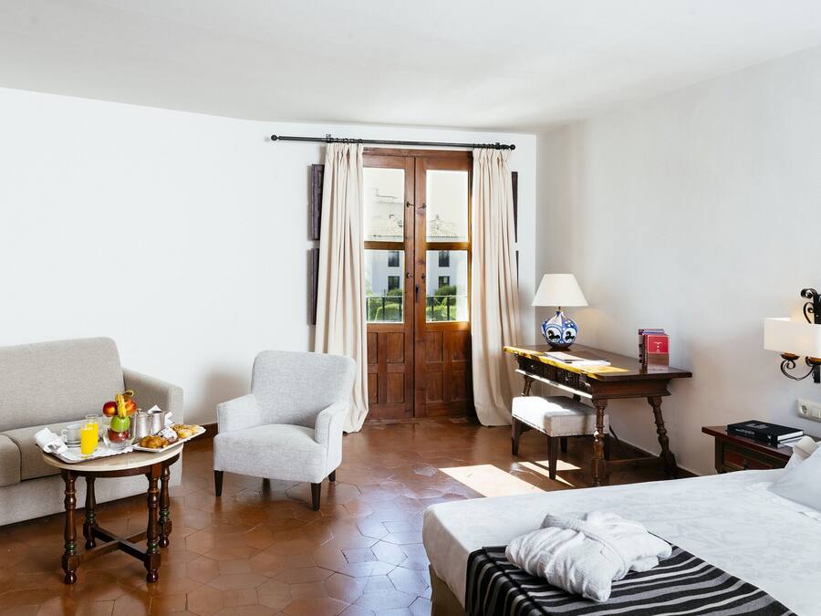 Quarto de hotel com cama, sofá, uma pequena mesa com café da manhã, escrivaninha e janela com cortinas.