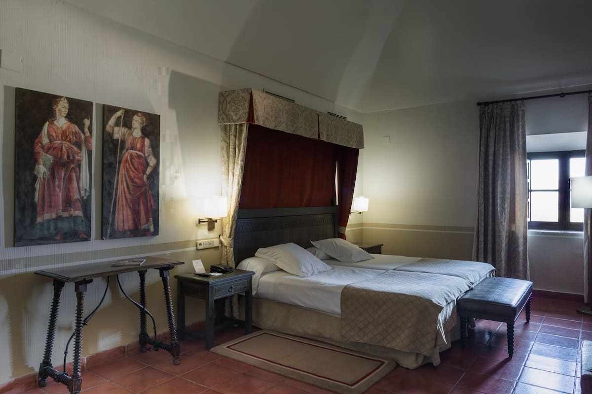 Quarto de hotel com uma cama grande, mesas de cabeceira, um banquinho e duas pinturas na parede.