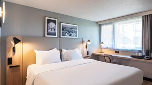 Quarto de hotel com uma cama, dois quadros pendurados na parede, uma mesa com abajur e uma janela com cortinas.