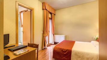 Quarto de hotel com uma cama, uma escrivaninha com um computador e um livro, uma cadeira e uma janela com cortinas.