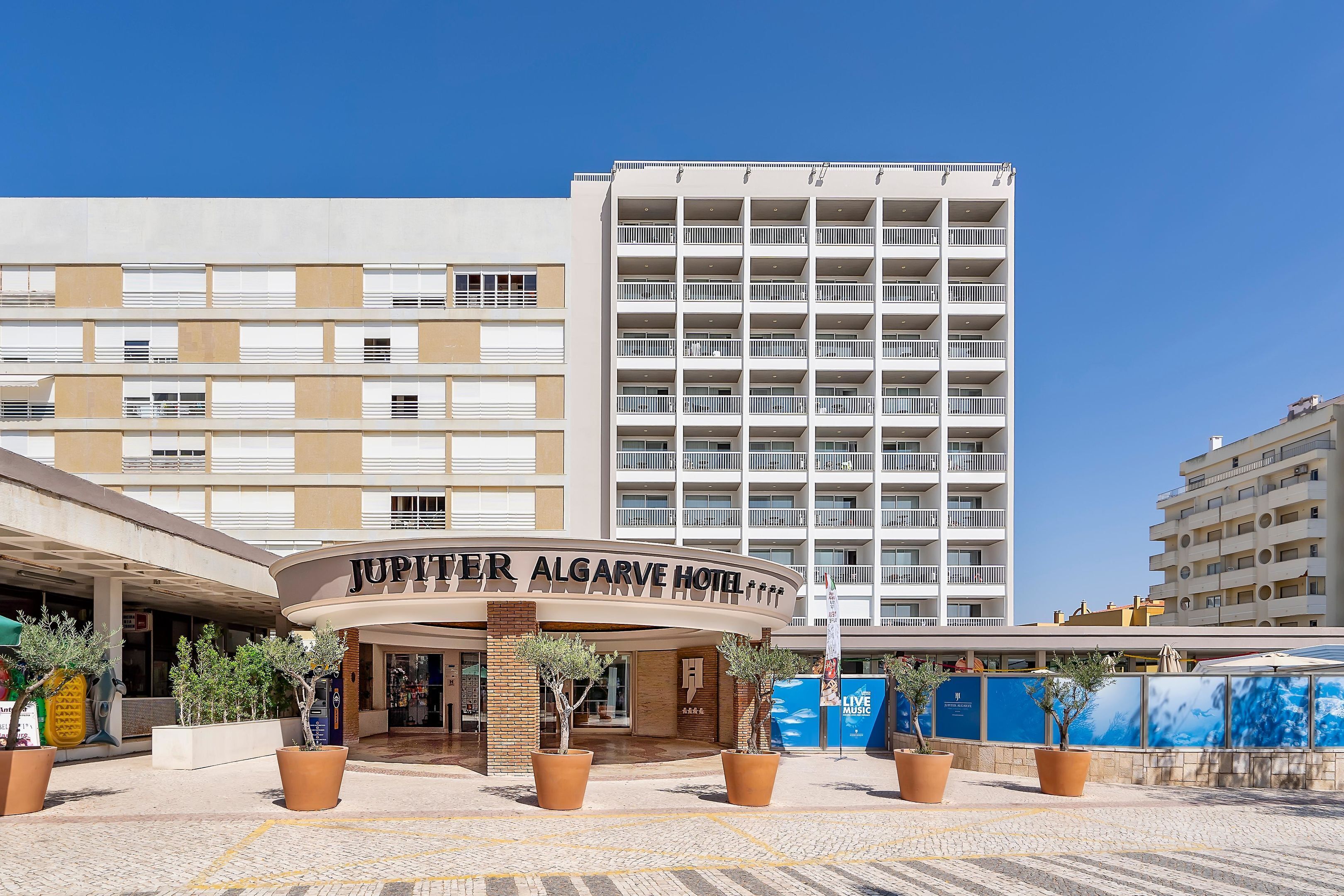 Entrada de hotel com fachada curva, várias varandas e plantas em vasos.