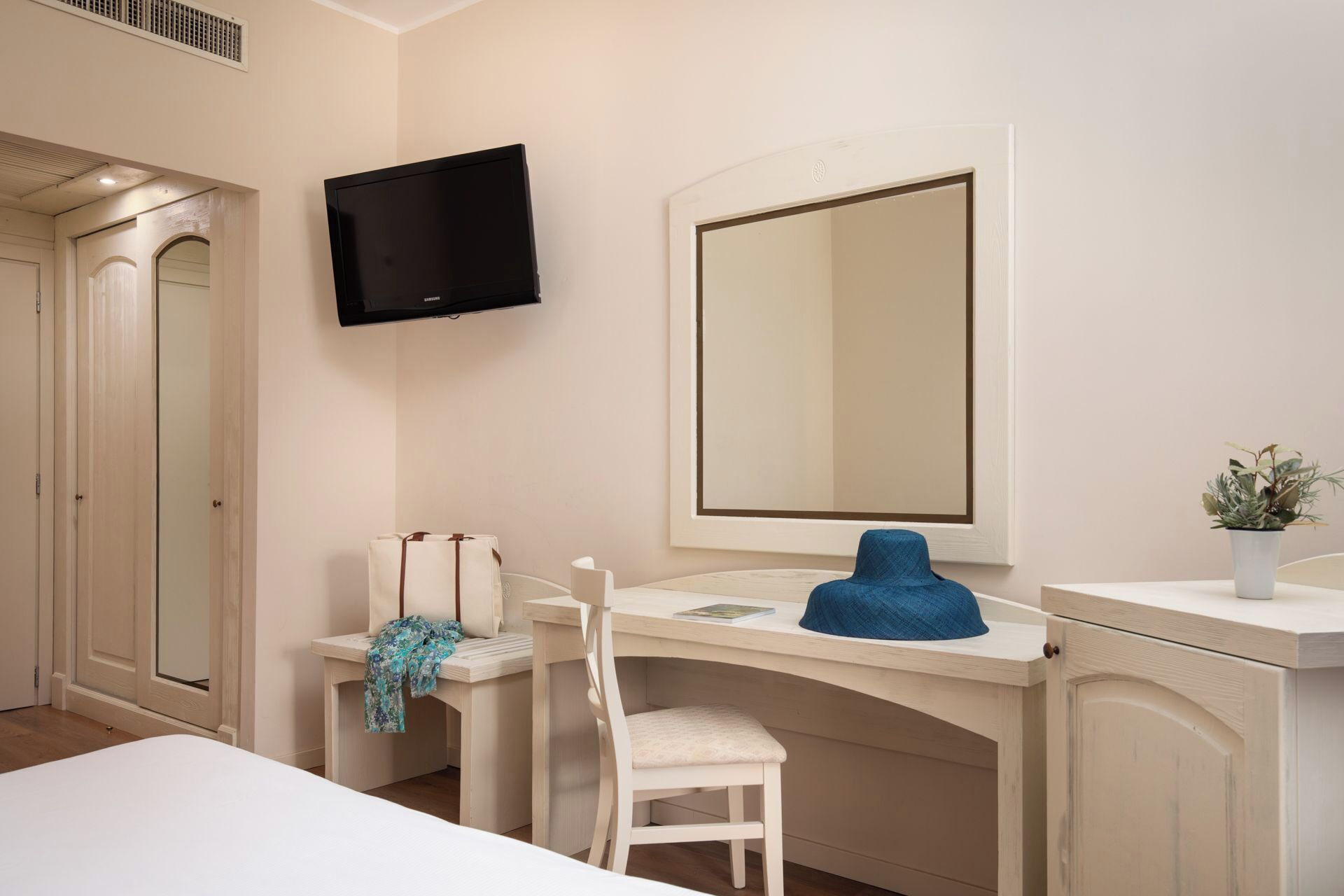 Quarto de hotel com uma cama, uma mesa com cadeira, televisão na parede, um espelho grande e um vaso de planta.