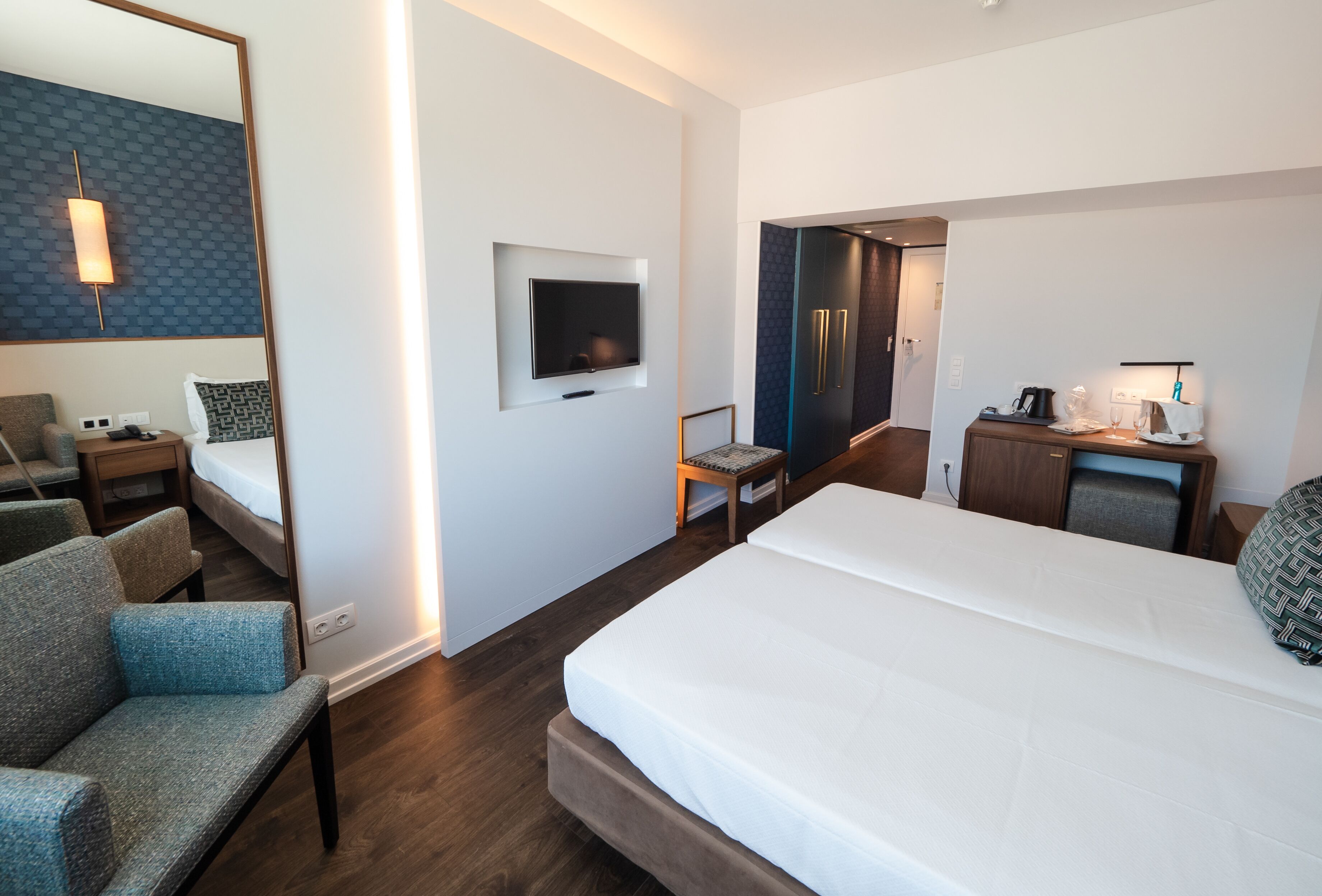 Quarto de hotel com uma cama grande, televisão na parede, escrivaninha com abajur e uma cadeira.