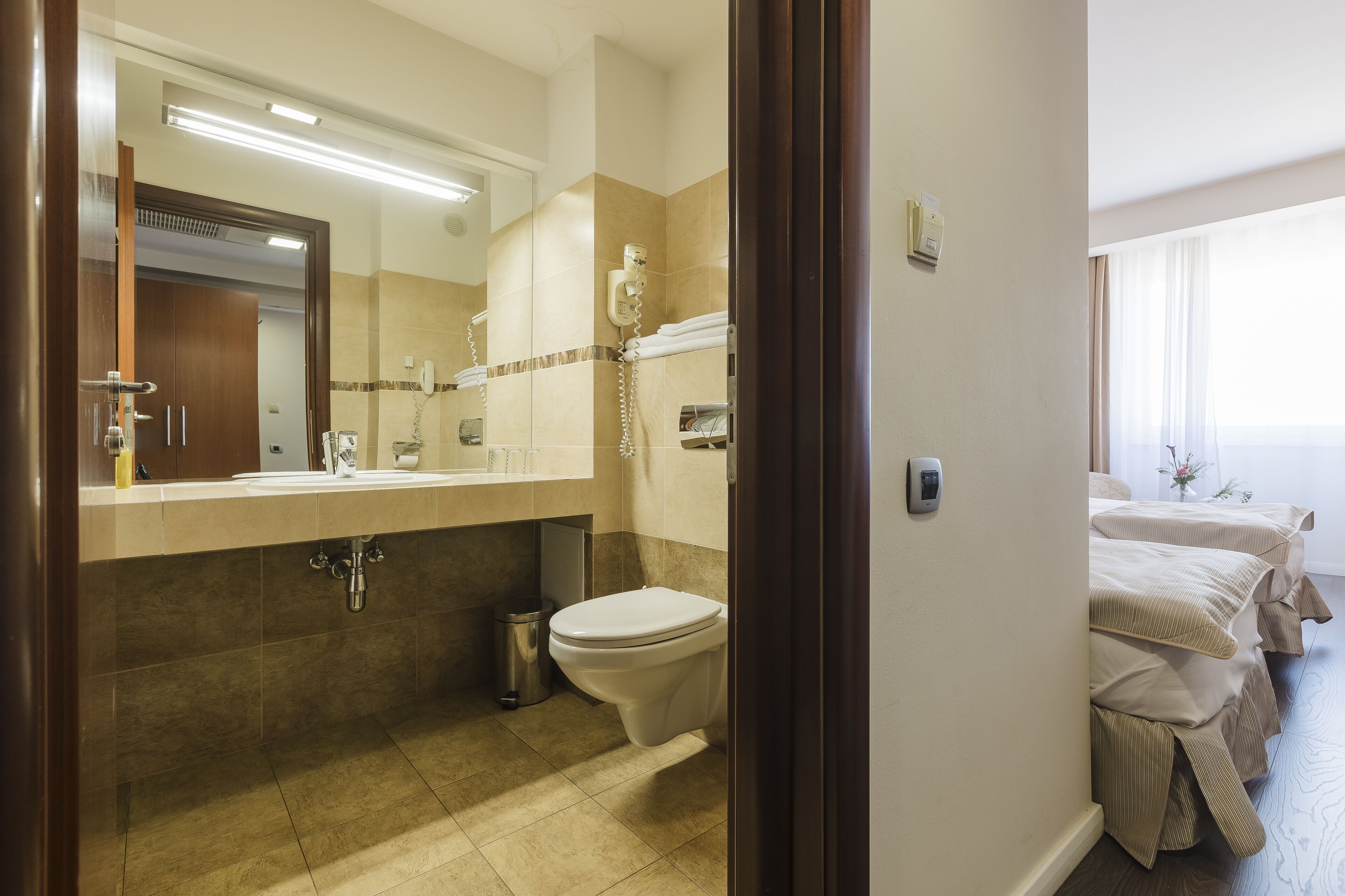 Casa de banho de hotel com sanita, lavatório e chuveiro. Há um contentor de lixo e um porta-toalhas.