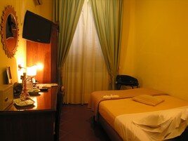 Quarto de hotel com cama, escrivaninha com luminária, televisão e janela com cortinas.