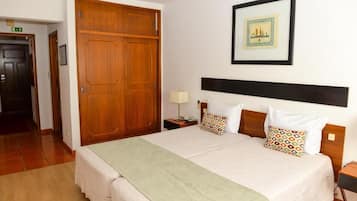 Quarto de hotel com uma cama grande, um guarda-roupa de madeira, mesa de cabeceira com abajur e um quadro na parede.