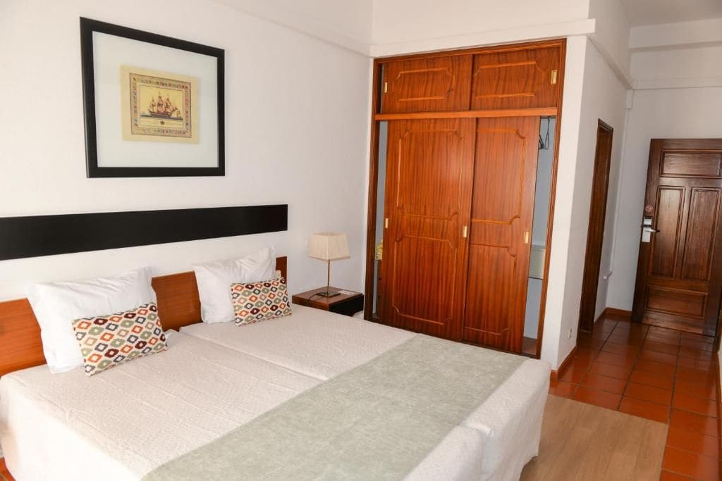 Quarto de hotel com cama grande, guarda-roupa de madeira, quadro emoldurado na parede e mesa de cabeceira com abajur.