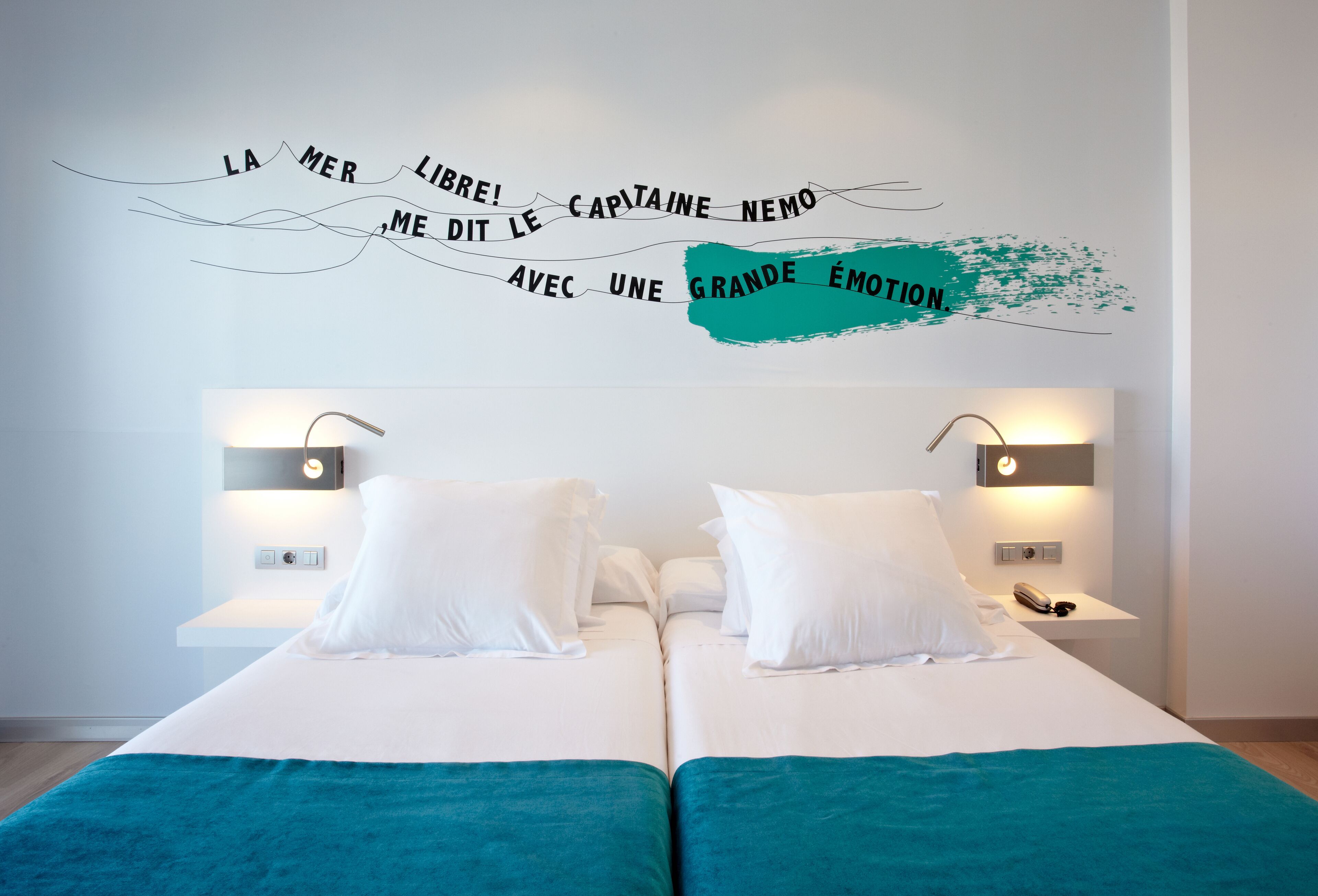 Quarto de hotel com duas camas, roupa de cama branca, mantas azuis e decoração na parede com texto em francês.