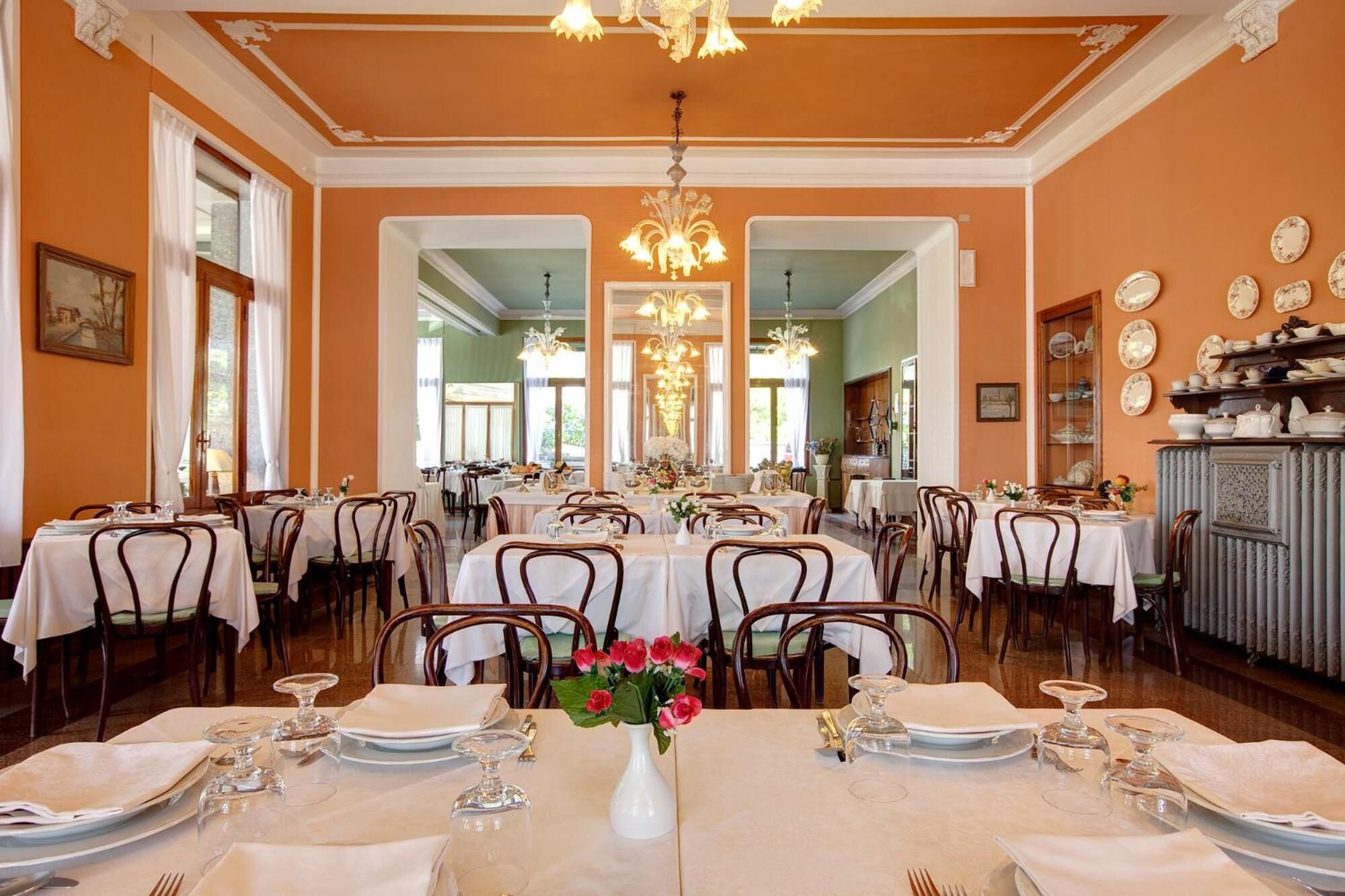 Uma sala de jantar com várias mesas preparadas para o serviço, um lustre e um vaso de flores.