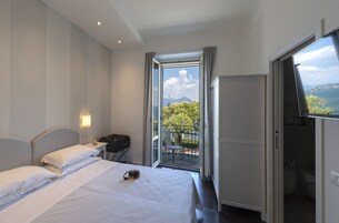 Quarto de hotel com cama, escrivaninha, cadeira e varanda com vista para o mar e montanhas.