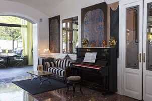 Um piano, um sofá e uma pequena mesa num quarto com paredes estampadas e uma porta com painéis de vidro decorativos.