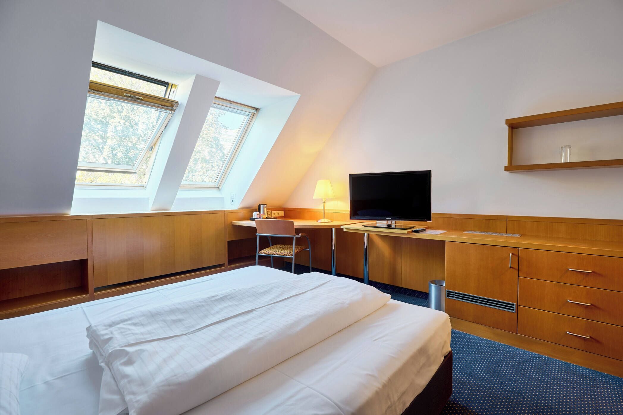 Quarto de hotel com cama, escrivaninha, cadeira, televisão e janela com vista para árvores.