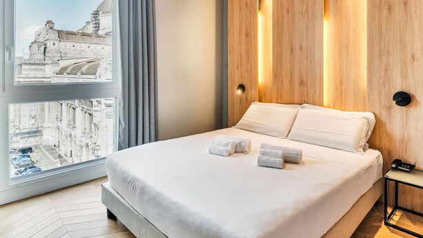 Quarto de hotel moderno com uma cama grande, duas mesas de cabeceira e uma janela ampla com vista para a cidade.